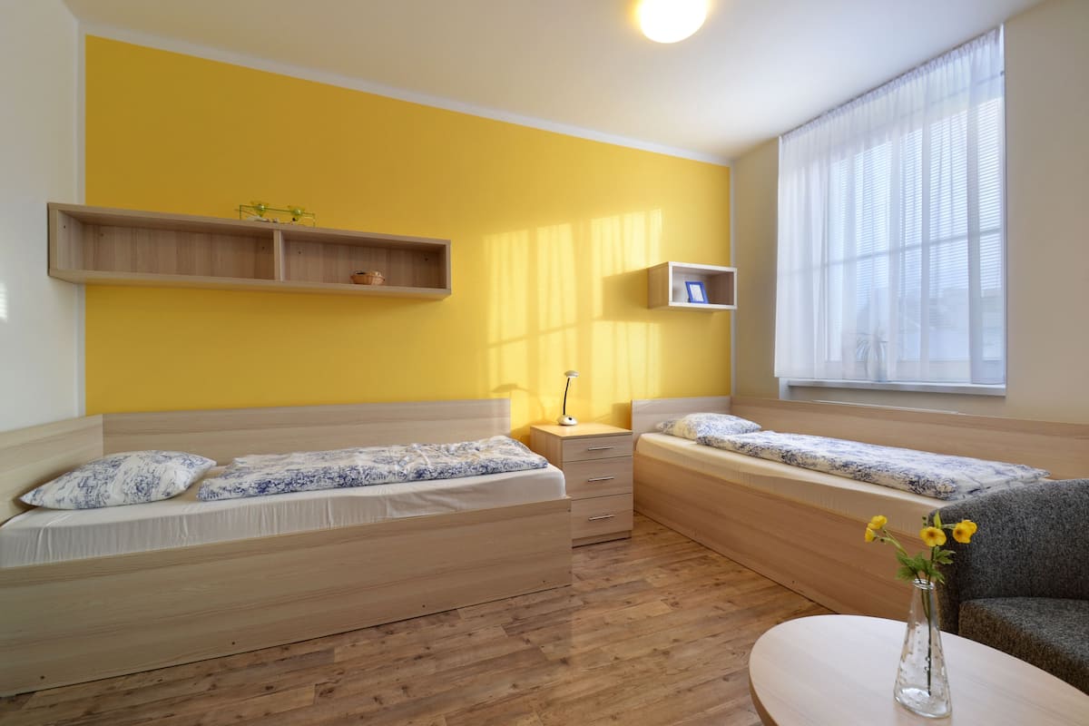 Quarto com duas camas, uma mesinha, um vaso de flores e uma janela com cortinas.