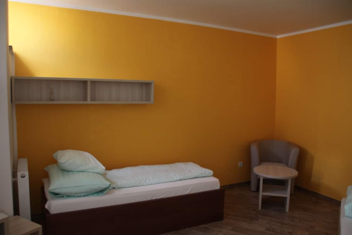 Quarto pequeno com cama de solteiro, cabeceira de madeira, uma cadeira e prateleira sobre a cama.
