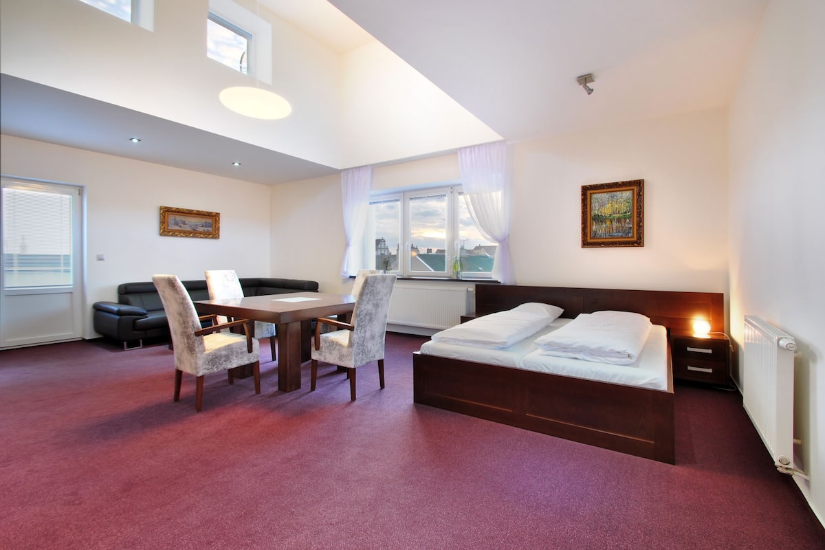 Quarto de hotel com cama, escrivaninha, duas cadeiras, janela com vista e um quadro na parede.