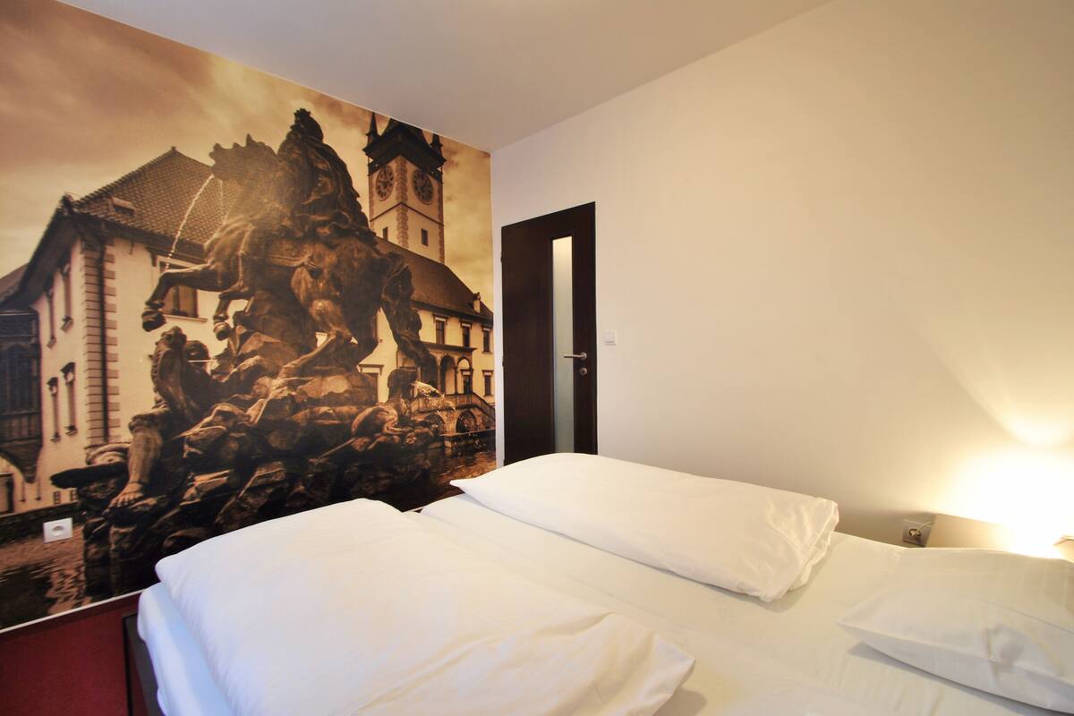 Quarto de hotel com um mural de um edifício histórico e uma cama com roupa de cama branca.