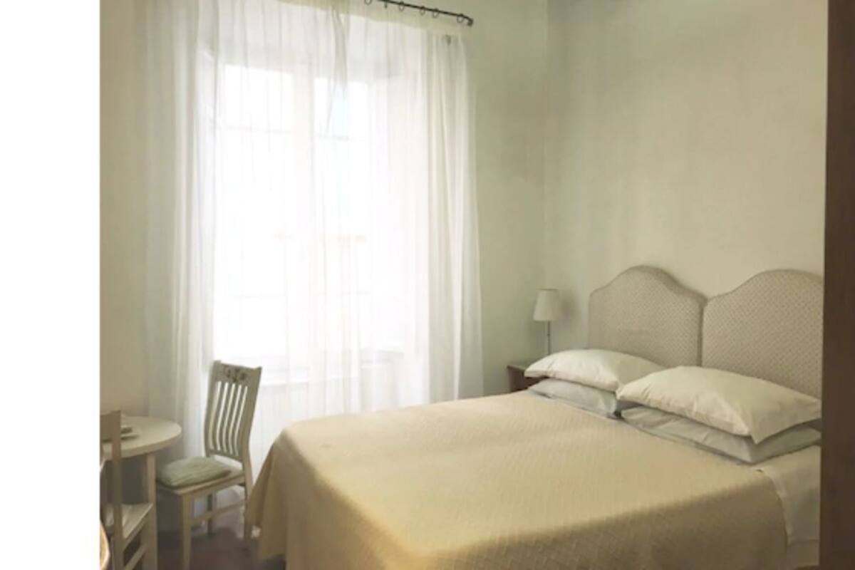 Quarto com cama, cadeira, mesa de cabeceira e janela com cortinas leves.
