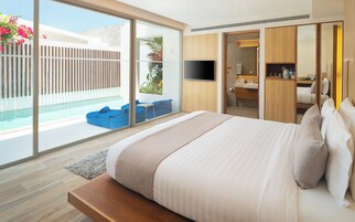 Quarto de hotel moderno com uma cama grande, uma TV, banheiro com pia e espelho, e varanda com piscina e cadeiras azuis.
