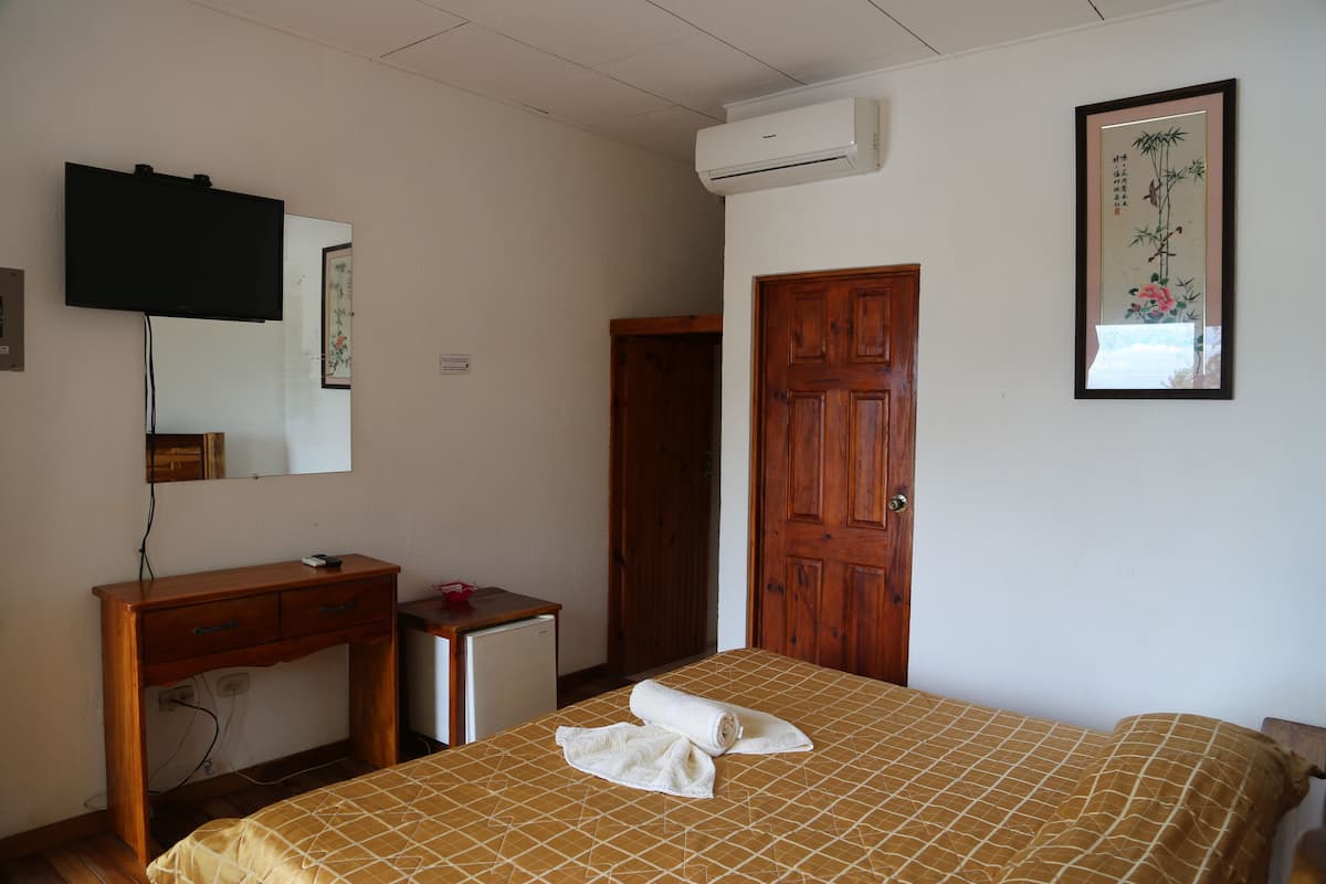 Quarto de hotel com uma cama, uma secretária de madeira, uma mesinha, um espelho, um ar condicionado e uma porta.