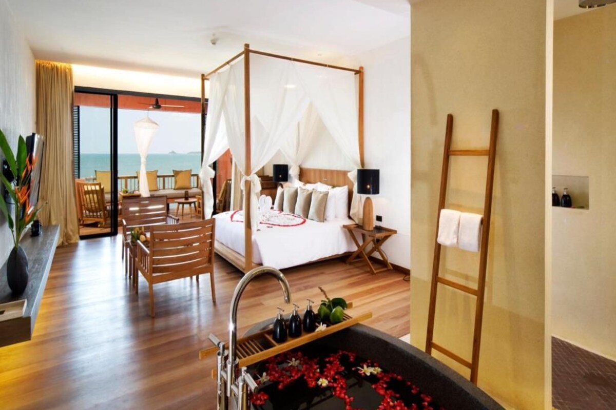 Um quarto espaçoso com cama de dossel, área de estar com mesa e cadeiras, janela grande com vista e banheira com pétalas de rosa.