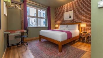 Quarto de hotel com uma cama, uma escrivaninha, uma cadeira, uma janela com cortinas, uma parede de tijolos, um quadro, um abajur e um tapete.