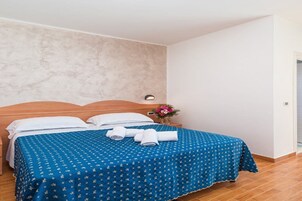 Uma cama bem arrumada com colcha azul e almofadas brancas, uma pequena mesa de cabeceira com um vaso de flores e uma parede com padrão texturizado.