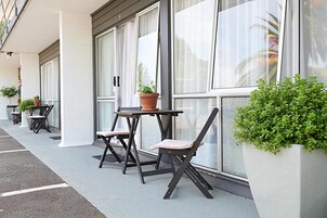 Exterior de hotel com área de estar coberta, plantas em vasos e uma grande janela.