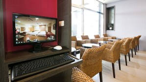 Quarto de hotel com escrivaninha, computador e uma grande janela. Há cadeiras de vime e uma mesa pequena no quarto.