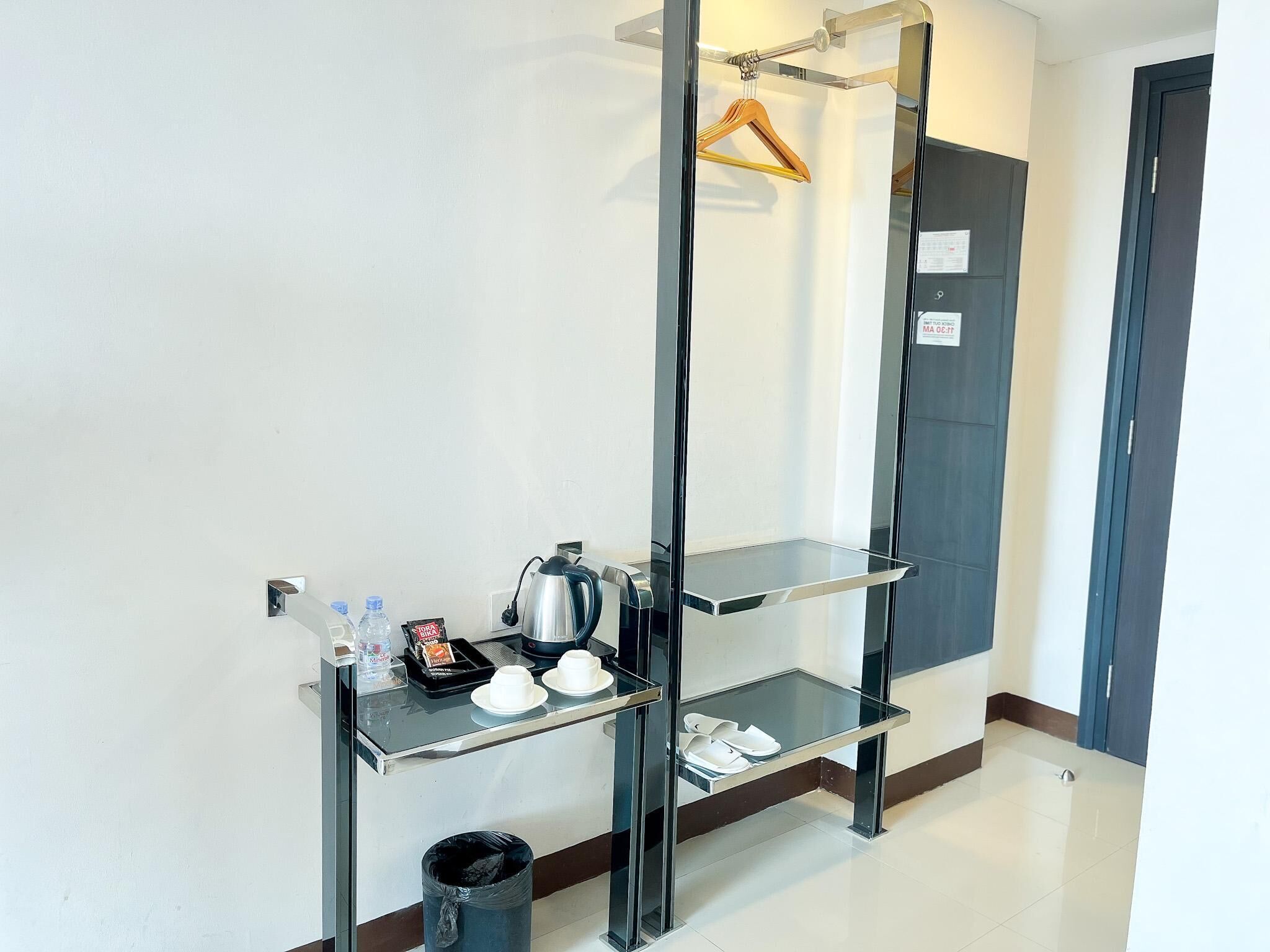 Quarto de hotel com divisória de vidro, uma mesinha com chaleira, chávenas e um prato com snacks. Há um cesto de lixo e um gancho para casacos na parede.