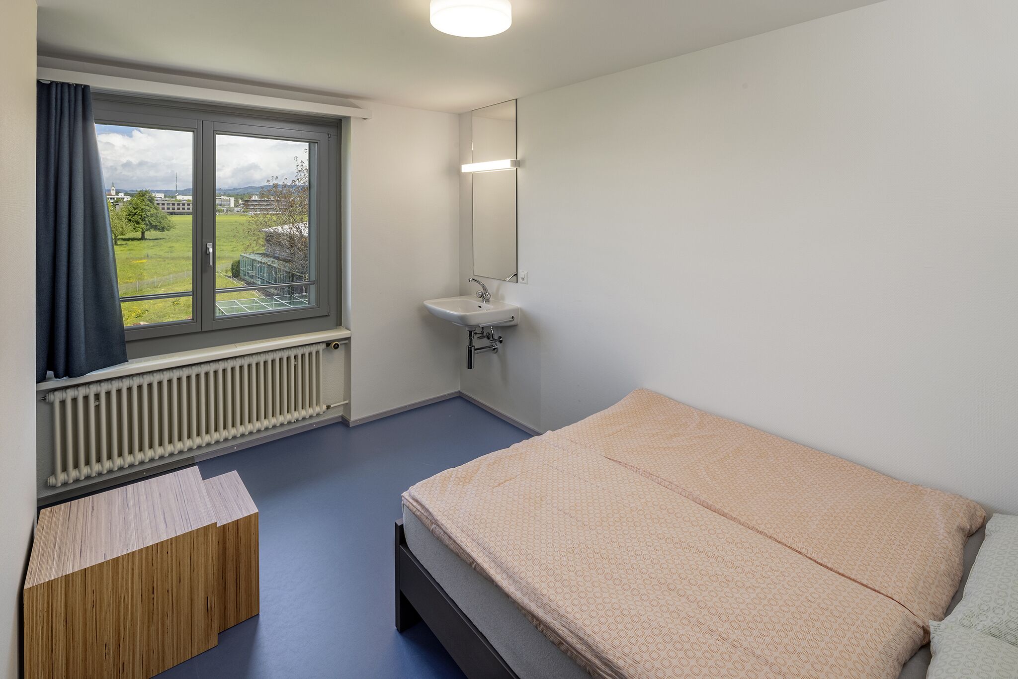 Quarto com cama, janela com vista para uma paisagem verde, pia de cor branca e radiador.