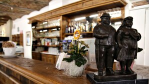 Uma área de bar com balcão de madeira, uma estátua de um homem e uma mulher, e prateleiras repletas de garrafas ao fundo.
