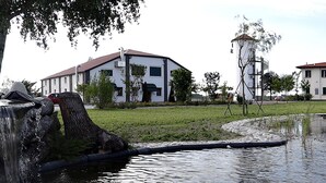 Um edifício com fachada branca e telhado vermelho, cercado por um gramado e um lago.