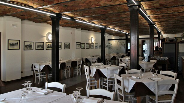 Um restaurante com mesas e cadeiras brancas, quadros nas paredes e tetos de tijolo aparente.