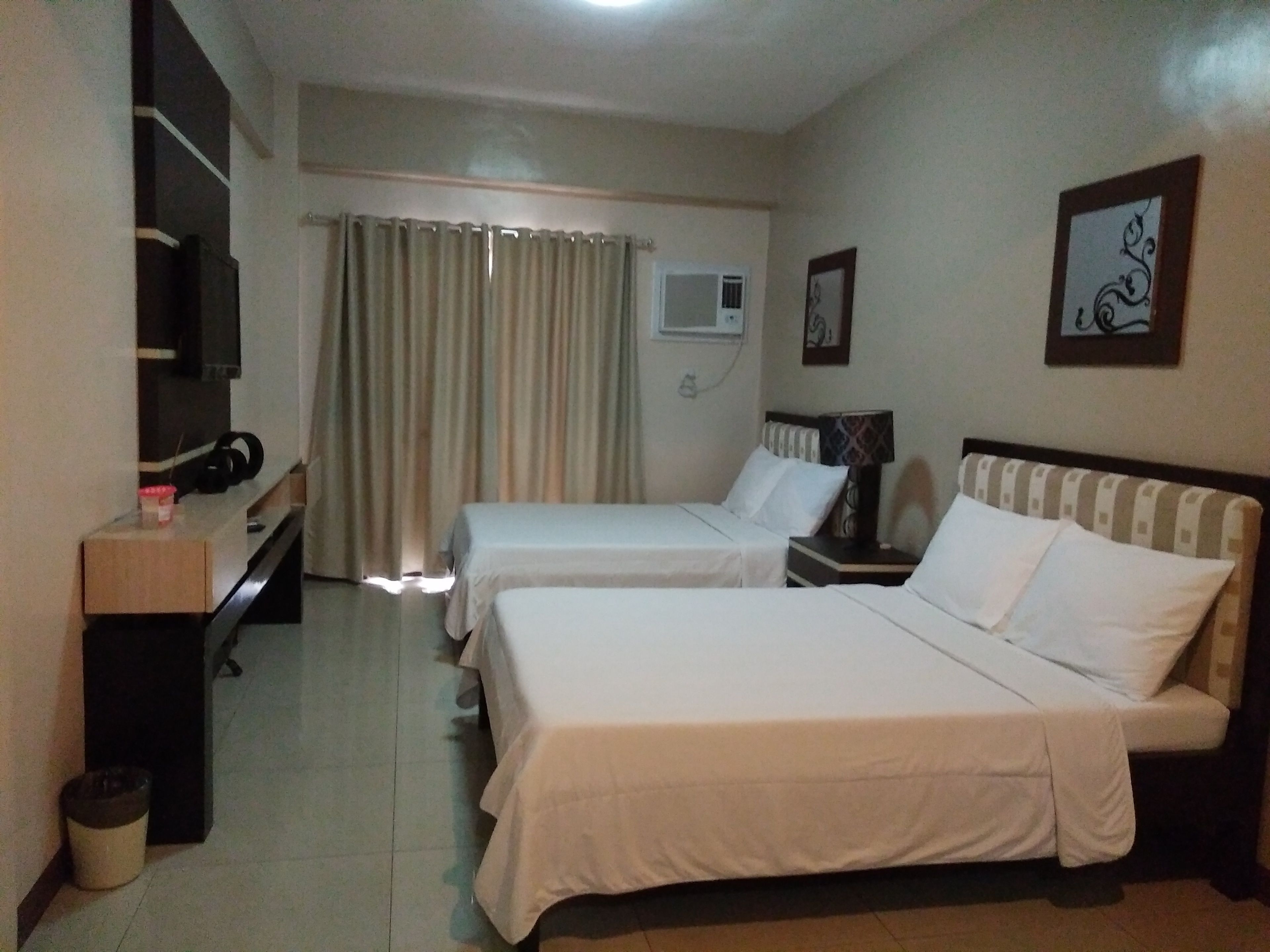 Quarto de hotel com duas camas, televisão, escrivaninha, lixeira e janela com cortinas.