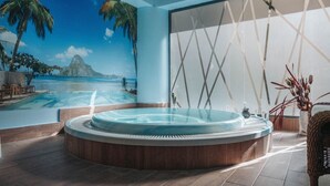 Uma banheira de hidromassagem em um quarto com um mural tropical, uma janela grande com persianas geométricas e uma cadeira de vime.