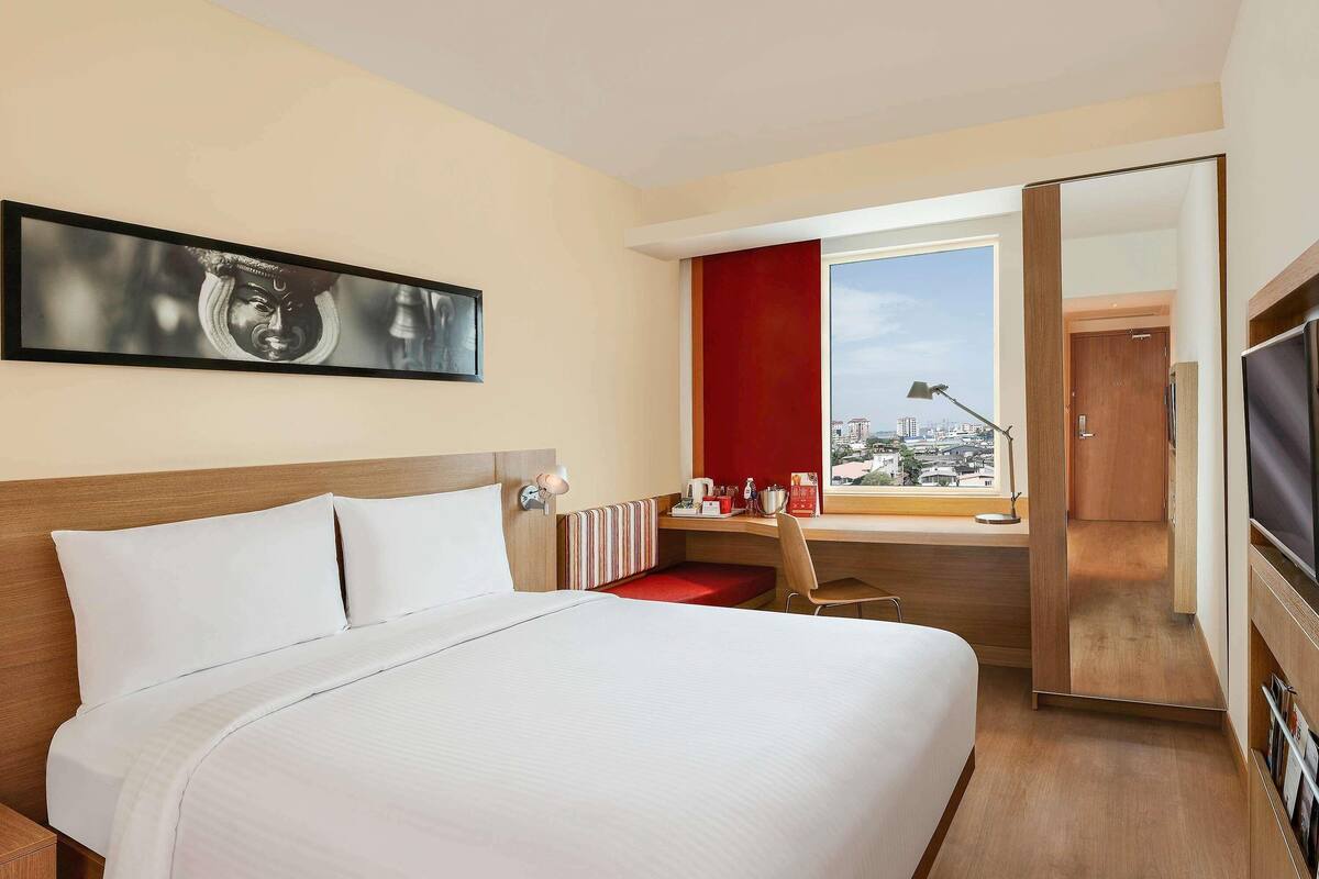 Quarto de hotel com uma cama grande, uma secretária com cadeira, janela com vista para a cidade e televisão.