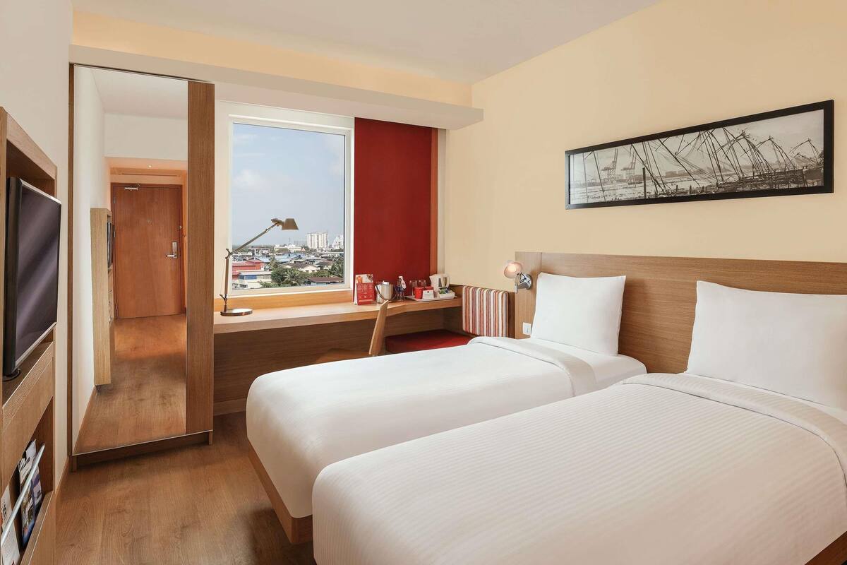 Quarto de hotel com duas camas, uma televisão, uma escrivaninha com candeeiro, uma janela com vista para a cidade e um quadro na parede.