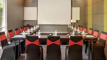 Sala de conferência com uma mesa comprida, cadeiras com fitas vermelhas, garrafas de água e maçãs sobre a mesa.