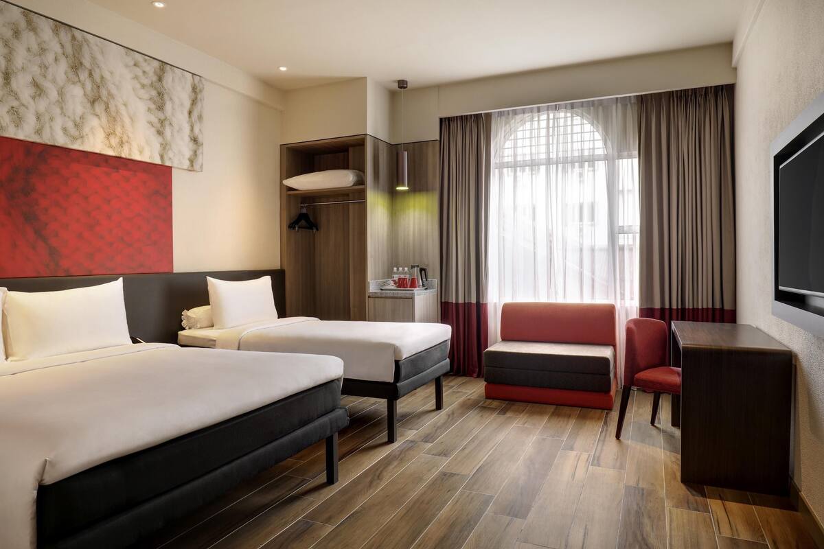Quarto de hotel com duas camas, uma escrivaninha, uma cadeira, um guarda-roupa e uma janela grande com cortinas.