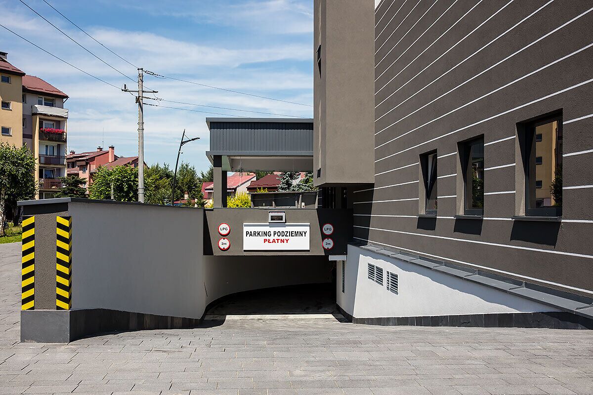 Um edifício moderno com entrada para estacionamento, um sinal indicando vagas disponíveis e uma área cercada com rampa de acesso à entrada.