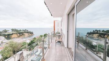 Varanda com guarda-corpo de vidro e vista para o litoral, mesa com cadeiras e céu limpo.