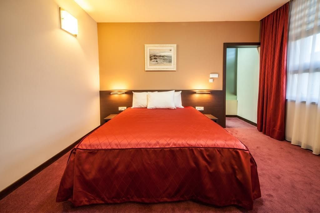 Quarto de hotel com uma cama grande, duas luminárias de cabeceira, um quadro na parede e vista pela porta.