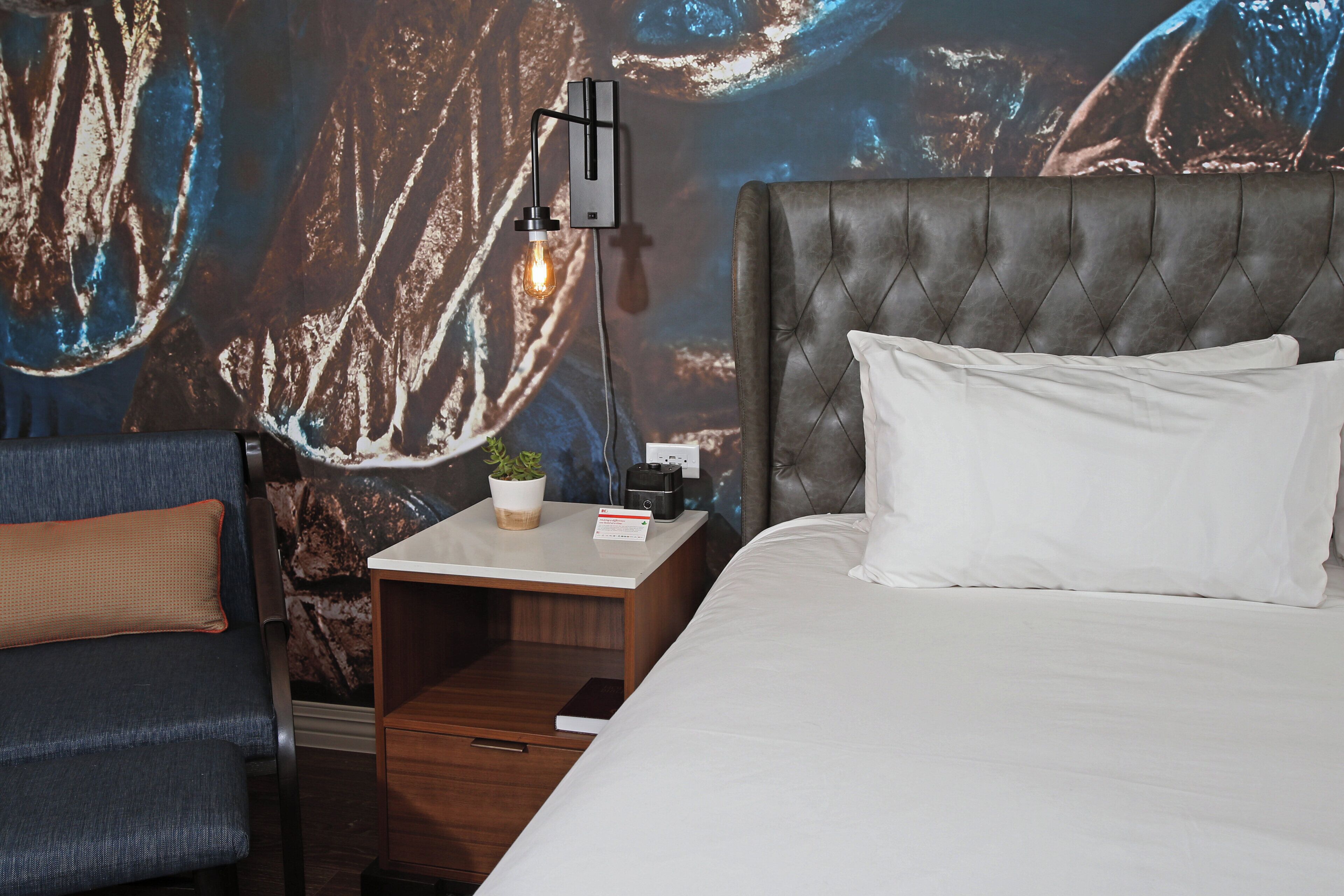 Quarto de hotel com cama, mesa de cabeceira, luminária, cadeira e um mural na parede.
