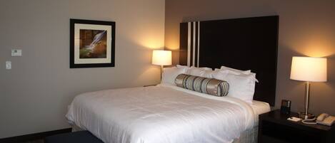 Quarto de hotel com uma cama grande, duas luminárias de cabeceira, mesa de cabeceira com telefone, quadro emoldurado na parede e um pequeno banco.