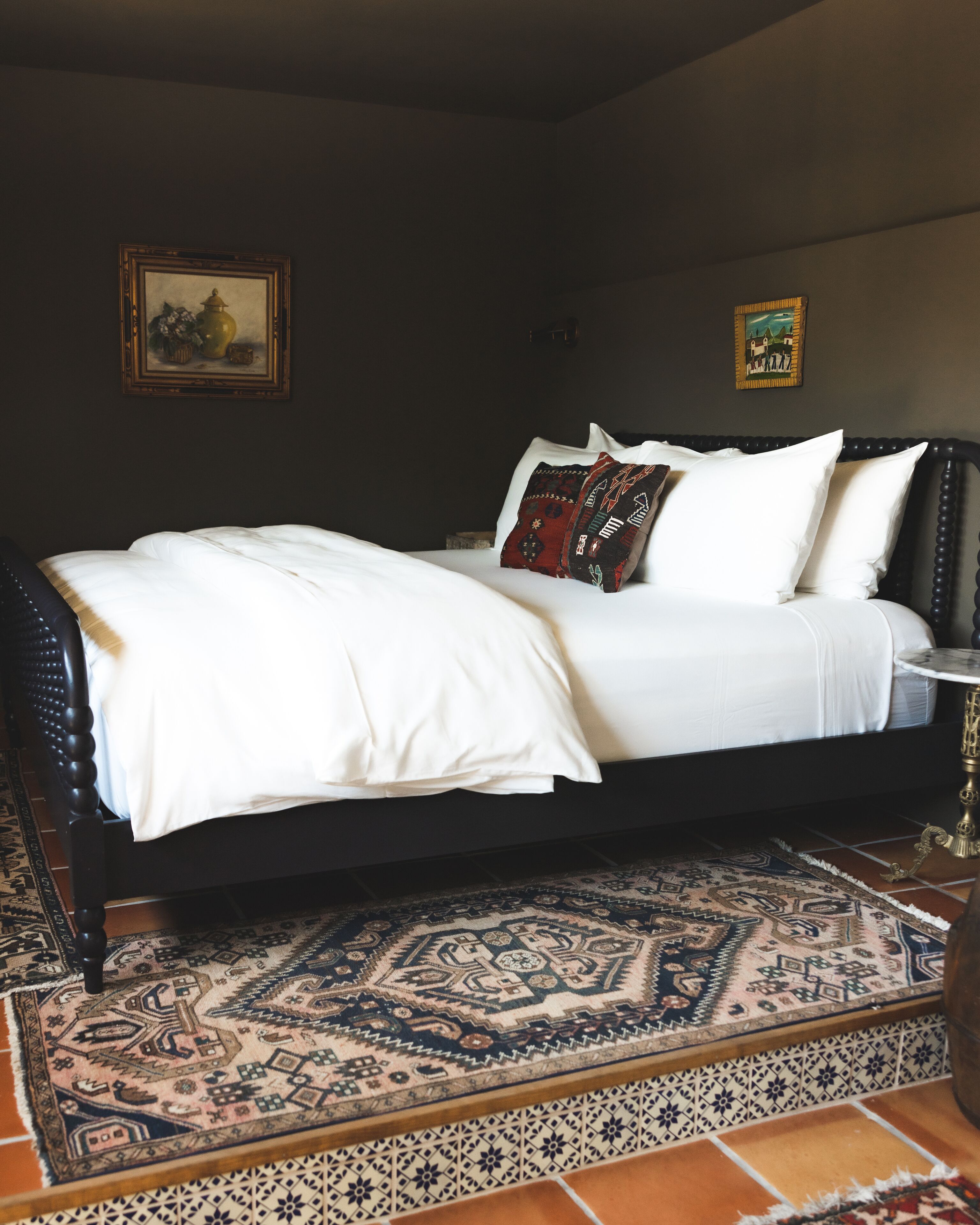 Uma cama com cabeceira escura, roupa de cama branca e um tapete estampado. Há dois travesseiros, um com padrão vermelho e preto.