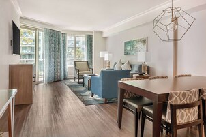 Quarto de hotel com piso de madeira, área de jantar com mesa e cadeiras, sofá, televisão fixada na parede e janela com cortinas.
