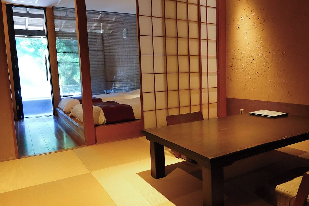 Quarto com mesa e cadeiras de madeira, porta deslizante que dá para uma cama com vista e um biombó shoji.