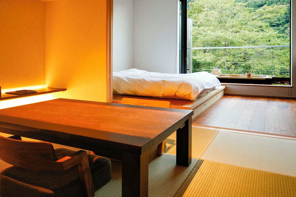 Quarto com cama, mesa e cadeira, e vista para o verde lá fora.
