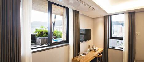Quarto de hotel com escrivaninha de madeira, televisão de tela plana e grandes janelas com vista para a cidade.