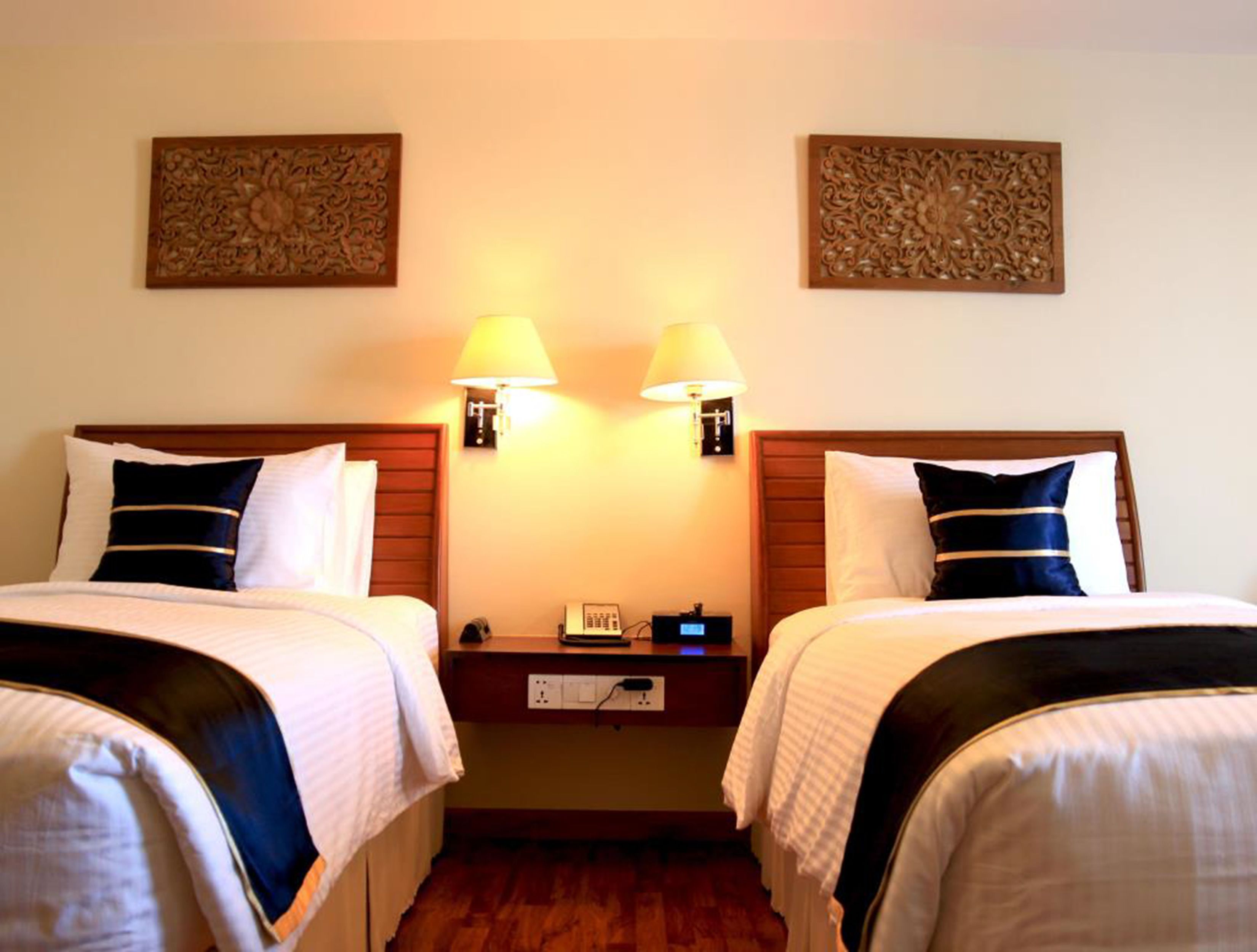Quarto de hotel com duas camas, cada uma com roupa de cama branca e detalhes em azul escuro, ladeadas por cabeceiras de madeira. Há duas luminárias na parede entre as camas, e uma pequena mesa com luminária e carregador de celular.