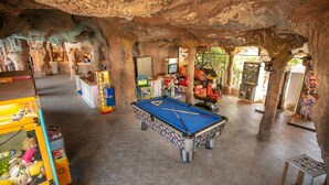 Uma mesa de bilhar numa sala que lembra uma caverna, com jogos de fliperama e área de bar.