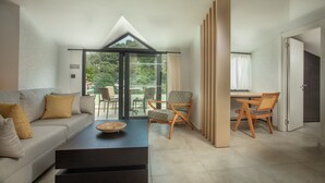 Uma sala de estar moderna com um sofá, mesa de centro e cadeiras. Há uma área de jantar com uma mesa e cadeiras. Uma grande janela oferece vista para o verde lá fora.
