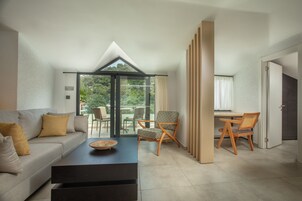 Uma sala de estar moderna com um sofá, mesa de centro e cadeiras. Há uma área de jantar com uma mesa e cadeiras. Uma grande janela oferece vista para o verde lá fora.