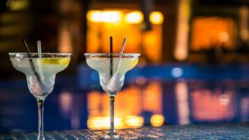 Dois copos de martini com canudos sobre uma superfície em mosaico perto de uma piscina.