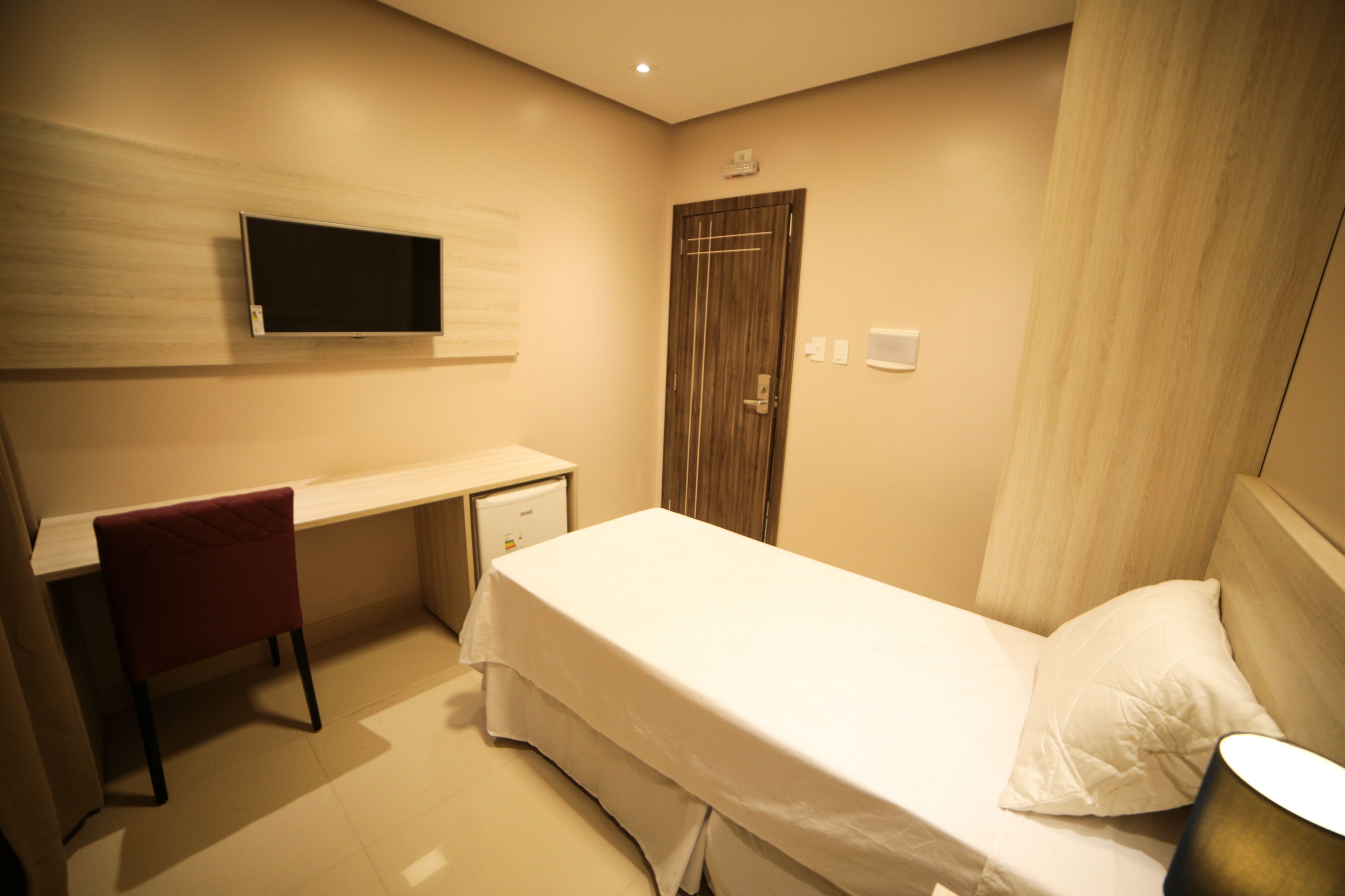 Quarto de hotel com uma cama de solteiro, uma mesa com cadeira, uma televisão fixada na parede e uma porta com puxador.