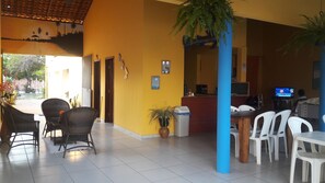 Um saguão de hotel com cadeiras de vime, um balcão de recepção, uma televisão e um vaso de plantas.
