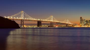 Vista noturna de uma grande ponte suspensa que atravessa um corpo de água, com luzes da cidade refletindo na superfície.