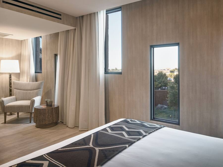 Quarto de hotel com uma cama grande, uma cadeira, uma mesa de apoio e vista para o exterior através de amplas janelas.