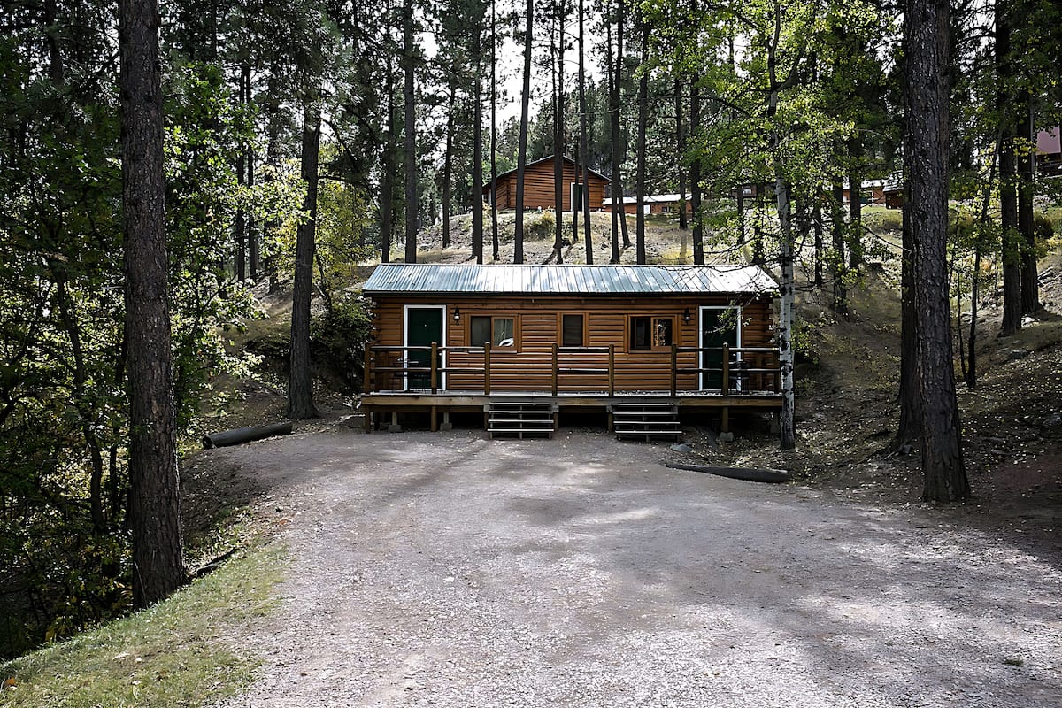 Uma cabana com telhado de metal, rodeada por árvores altas e um caminho de cascalho que a leva.