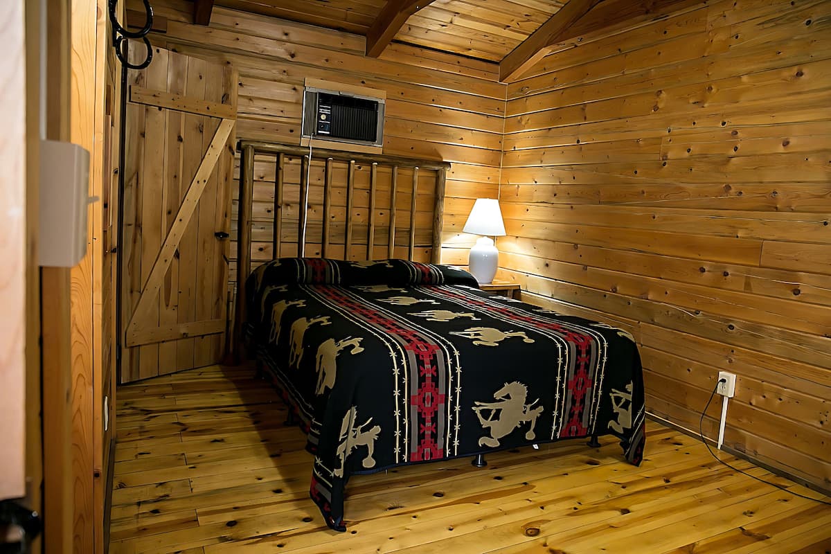 Quarto de cabana de madeira com uma cama, uma mesa de cabeceira, uma luminária e teto de madeira.