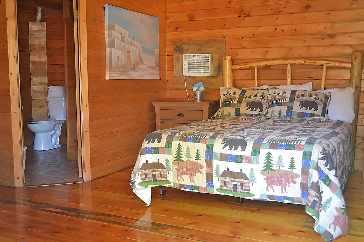 Quarto de cabana de madeira com uma cama, uma mesinha de cabeceira, um quadro e um toalete visível através de uma porta aberta.