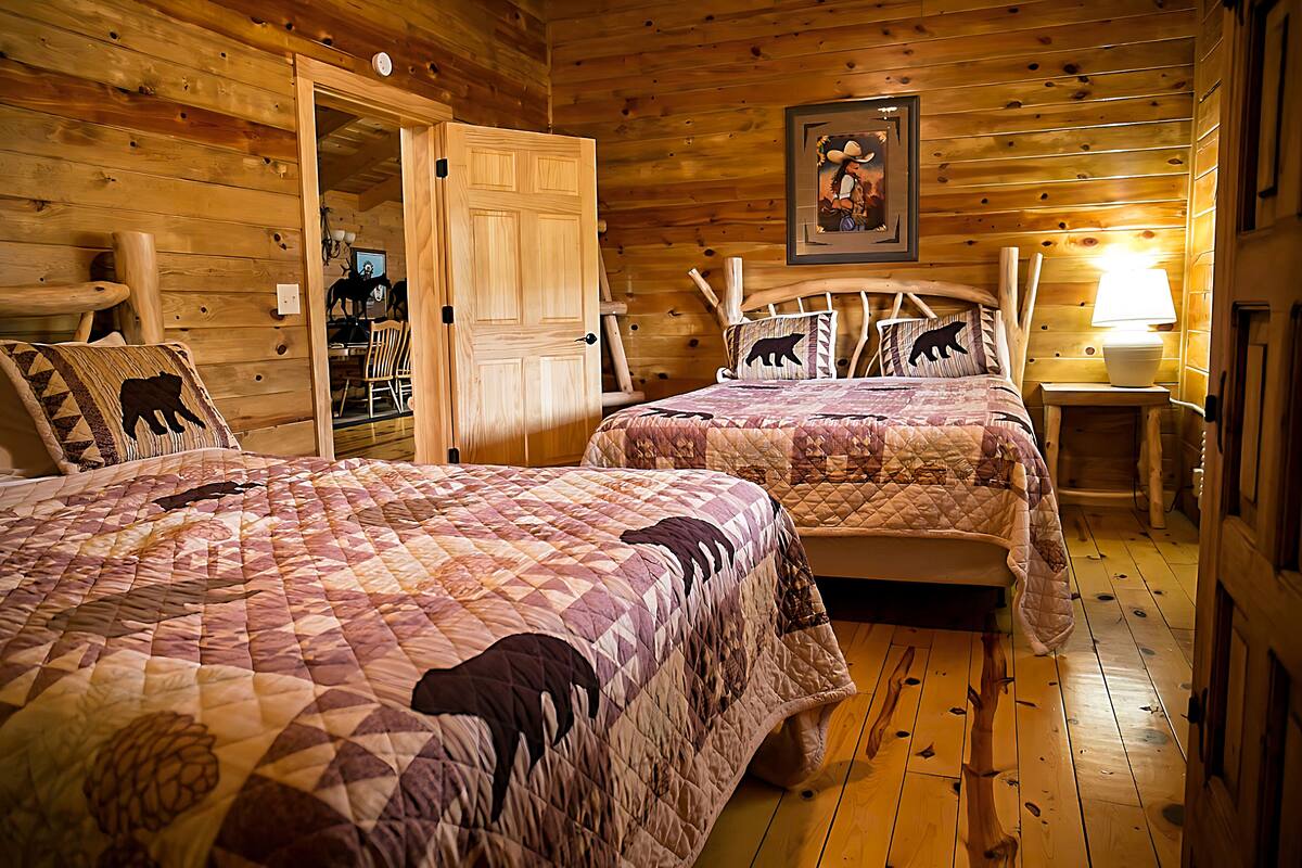 Um quarto estilo cabana de madeira com duas camas, uma porta de madeira, uma moldura com imagem na parede e uma pequena mesa com abajur.