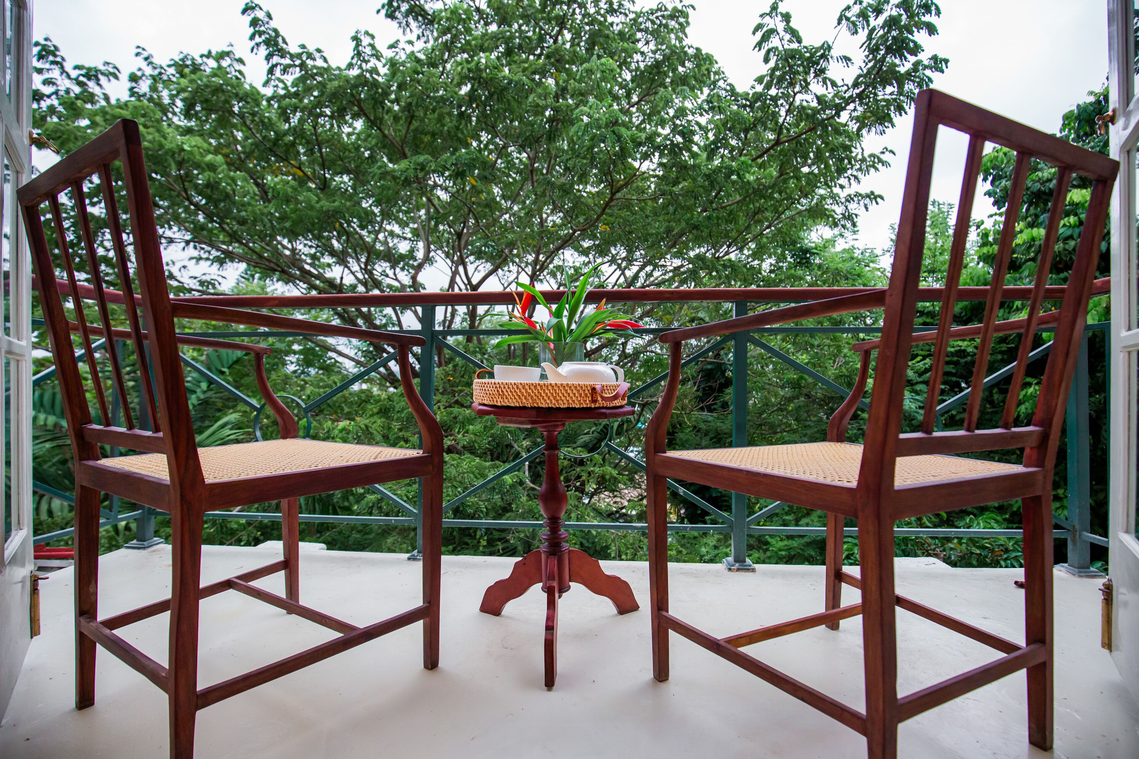 Duas cadeiras de madeira com assentos trançados e uma pequena mesa com uma cesta de vime, dispostas numa varanda com vista para árvores verdes.