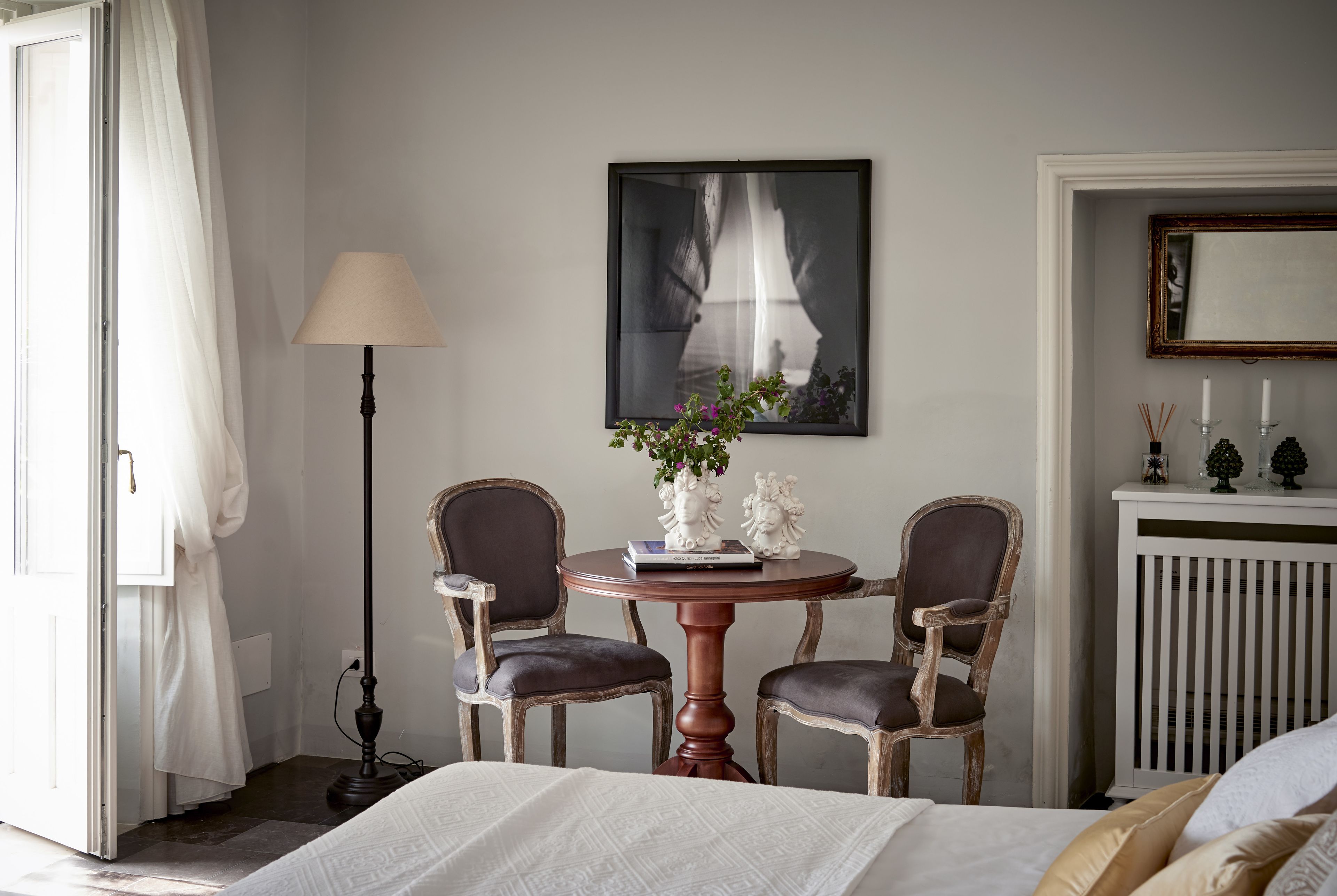 Quarto com cama, mesa redonda e cadeiras, abajur, quadro na parede e console branco com velas.