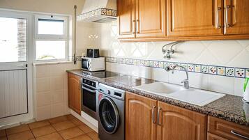 Cozinha compacta com armários de madeira, máquina de lavar embutida, micro-ondas e pia com painel de azulejos.