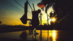 Vista do pôr do sol com uma taça de cocktail sobre uma mesa, palmeiras e uma praia ao fundo.